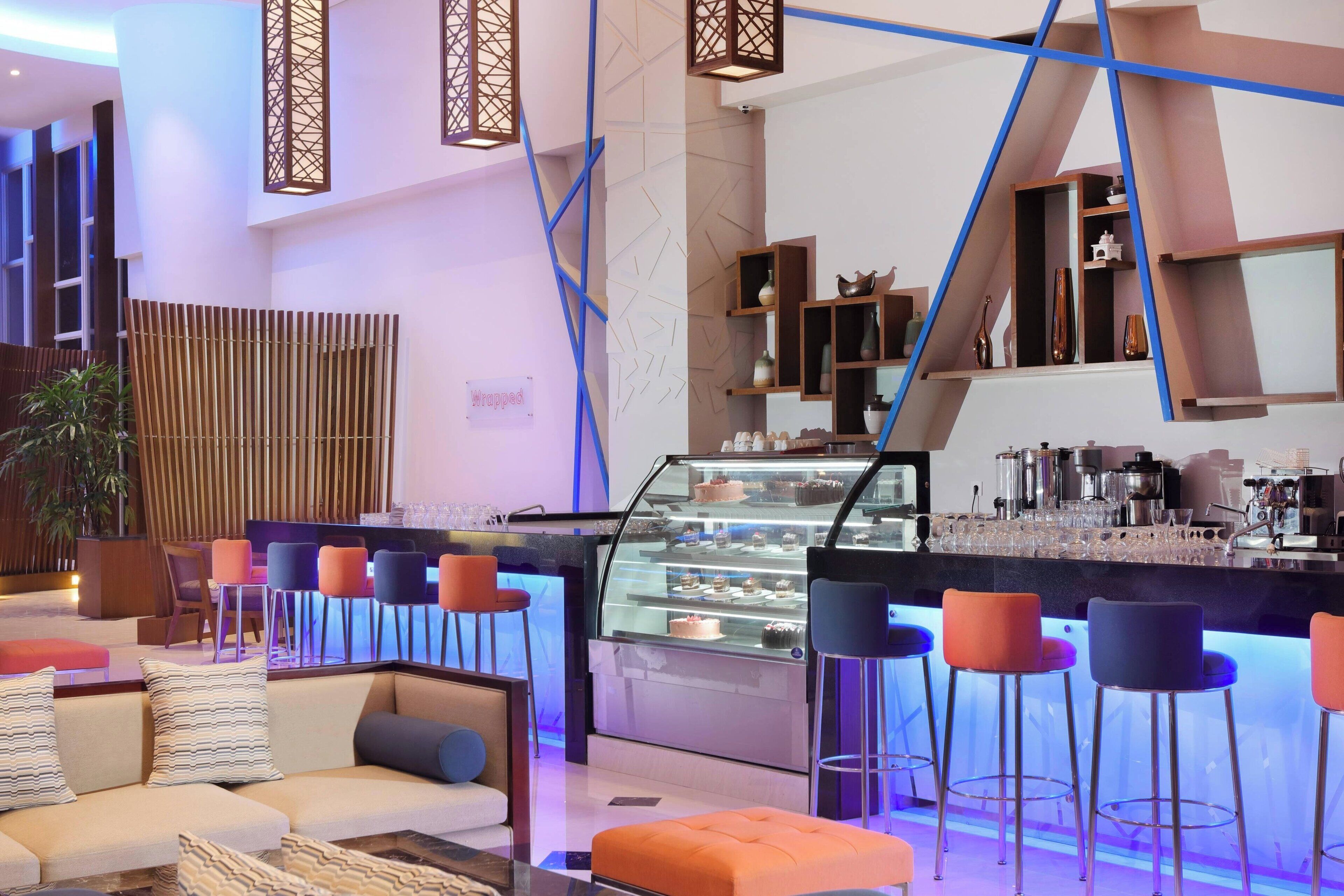 Uma área de bar moderna, com banquetas, uma vitrine de vidro com sobremesas e um espaço de estar com sofás e pufes.