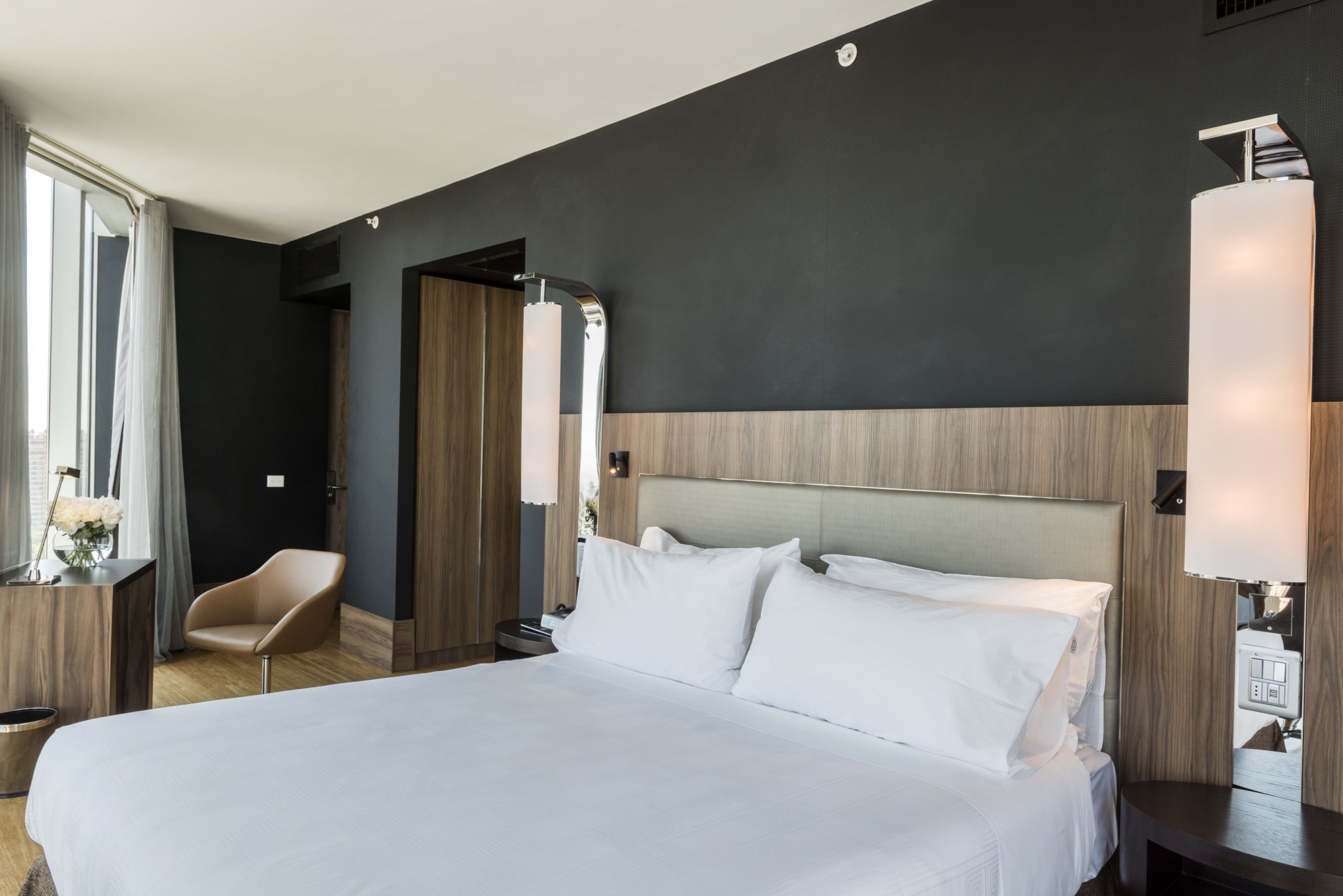 Quarto de hotel moderno com uma cama grande, cabeceira de madeira, escrivaninha com luminária e uma cadeira.