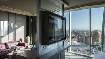 Uma sala de estar moderna com uma televisão de tela plana grande, um sofá de couro rosa, um vaso com flores e vista para o horizonte urbano através de janelas amplas.