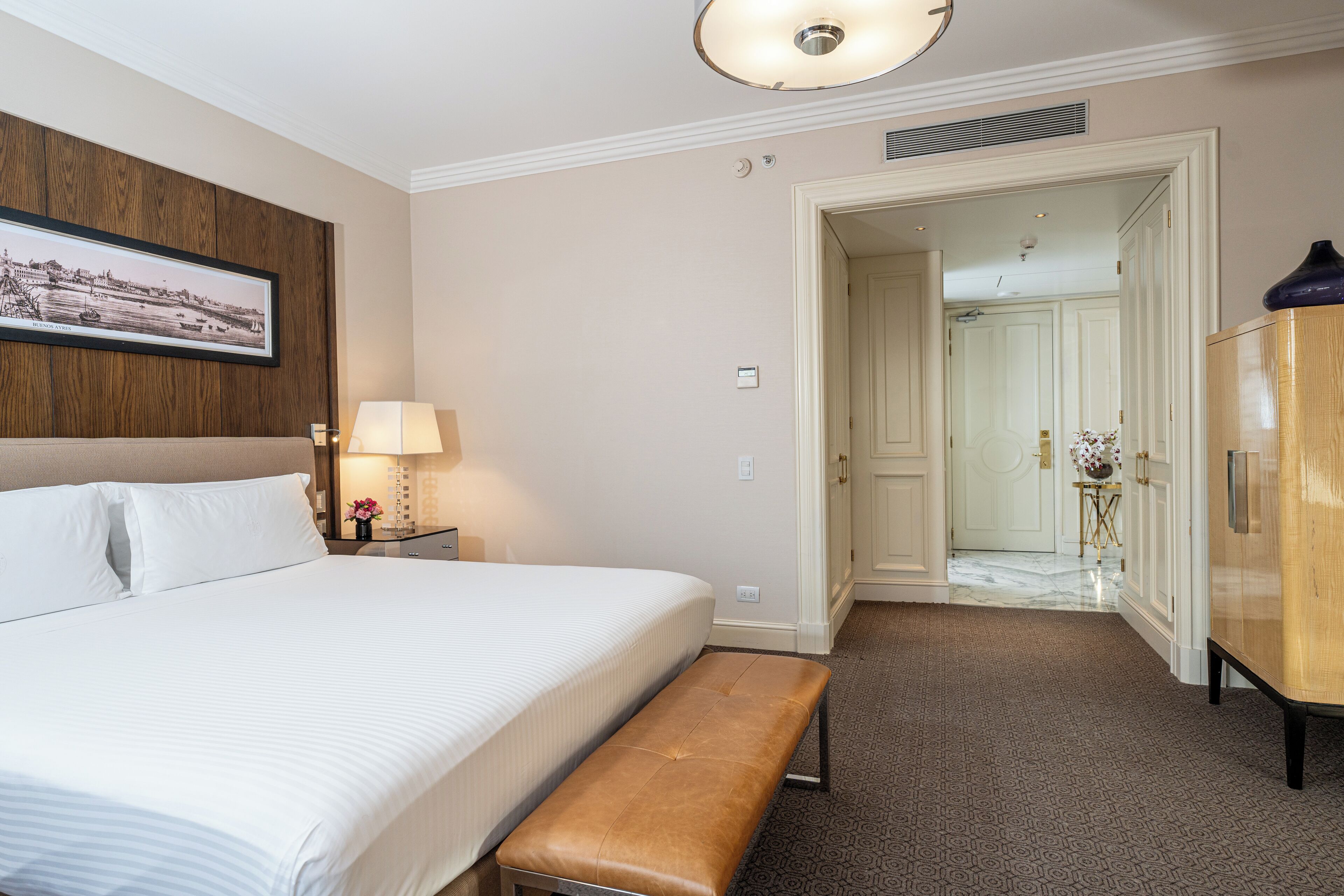 Quarto de hotel com uma cama grande, um banco, uma mesa de cabeceira com abajur, um quadro na parede e um guarda-roupa.