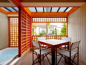 Um deck de madeira com mesa e cadeiras, uma banheira de hidromassagem e vista para uma casa através de uma treliça de madeira.