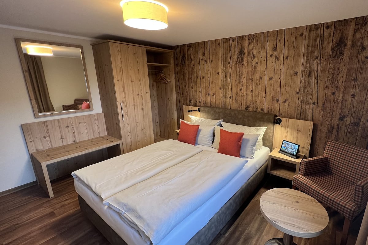 Quarto com cabeceira de madeira, cama com roupa de cama branca, mesa de cabeceira com um tablet, um banco e um espelho.
