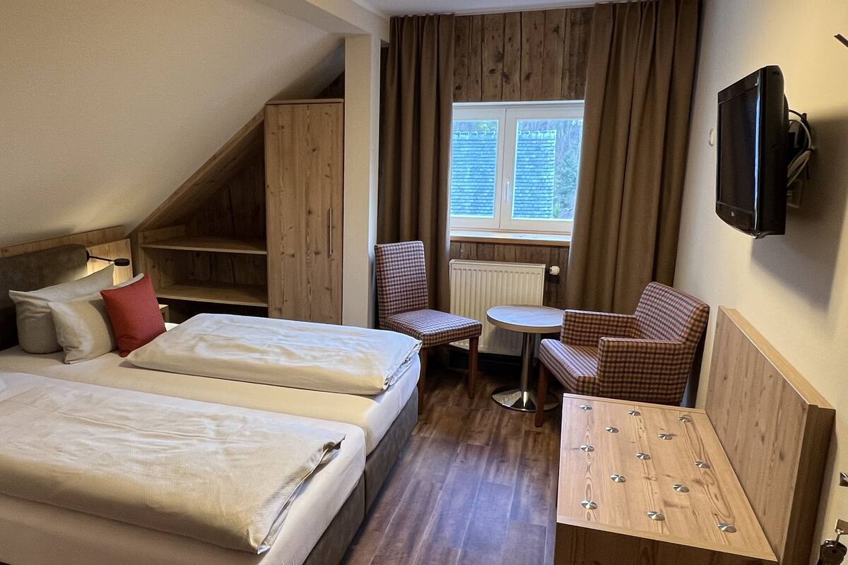 Quarto de hotel com duas camas, uma mesa pequena, uma cadeira, televisão e janela com cortinas.