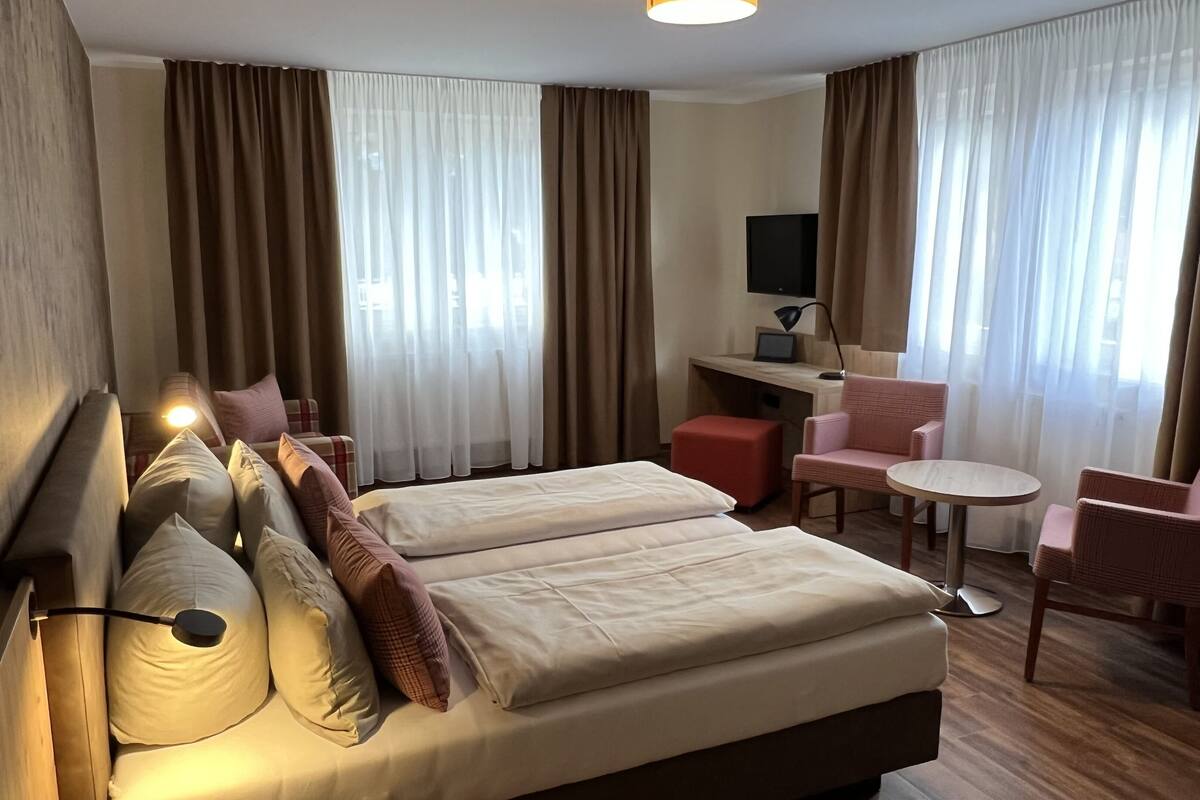 Quarto de hotel com uma cama de casal, uma escrivaninha com cadeira, uma mesa redonda pequena e uma televisão.