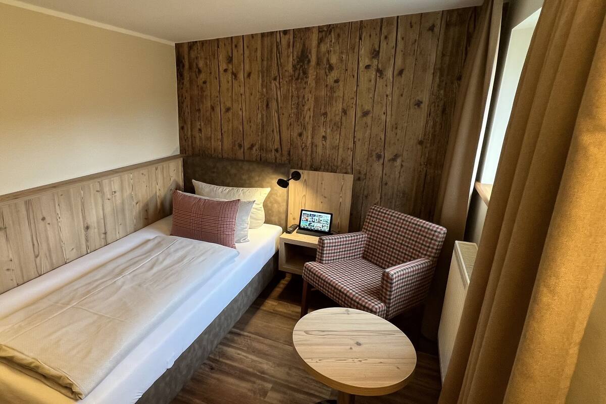 Quarto de hotel com uma cama, uma secretária pequena com um laptop, uma cadeira e uma mesa redonda.