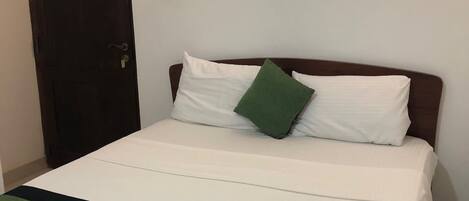 Cama bem arrumada com roupa de cama branca e verde, cabeceira de madeira escura e luminária fixada na parede.