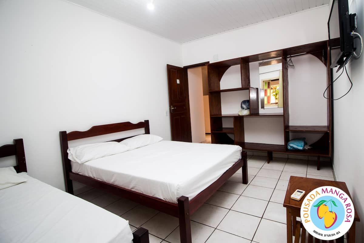 Quarto com uma cama de solteiro, um guarda-roupa de madeira, uma pequena mesa com um livro e uma televisão fixada na parede.
