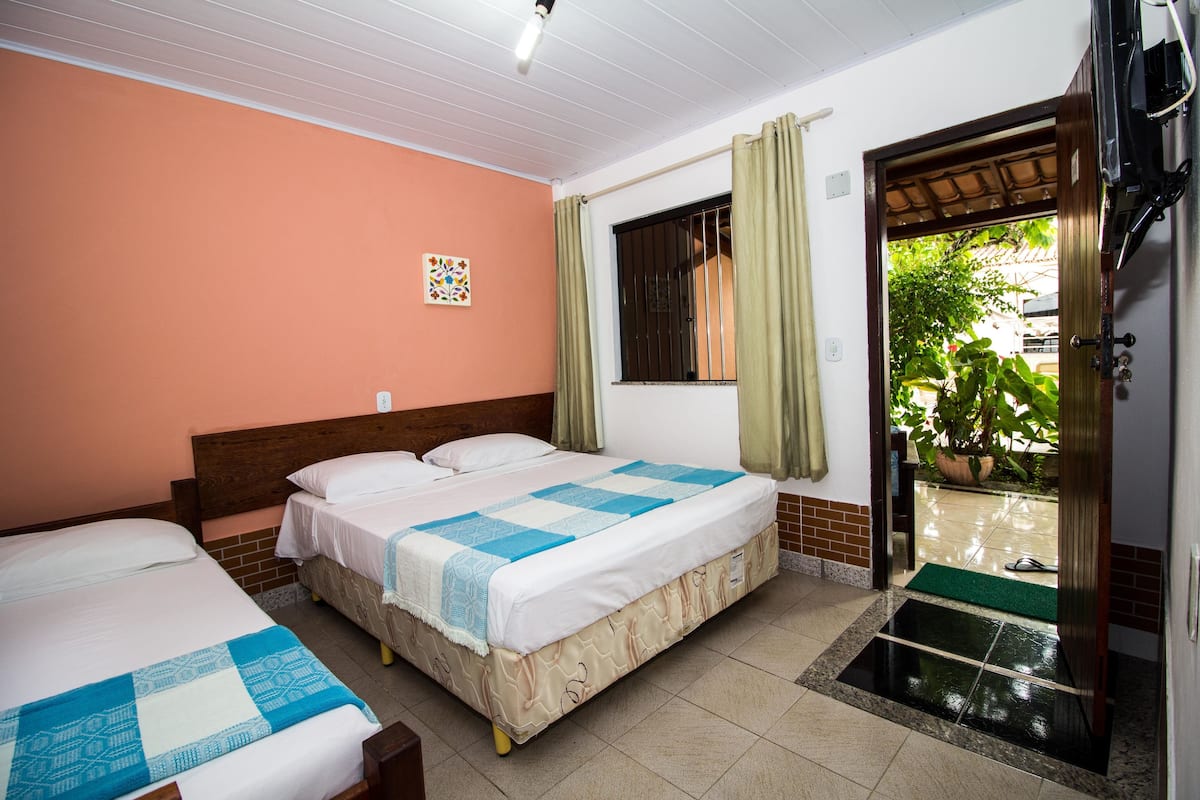 Quarto de hotel com duas camas, janela com cortinas, porta para o exterior e uma pequena varanda com planta em vaso.