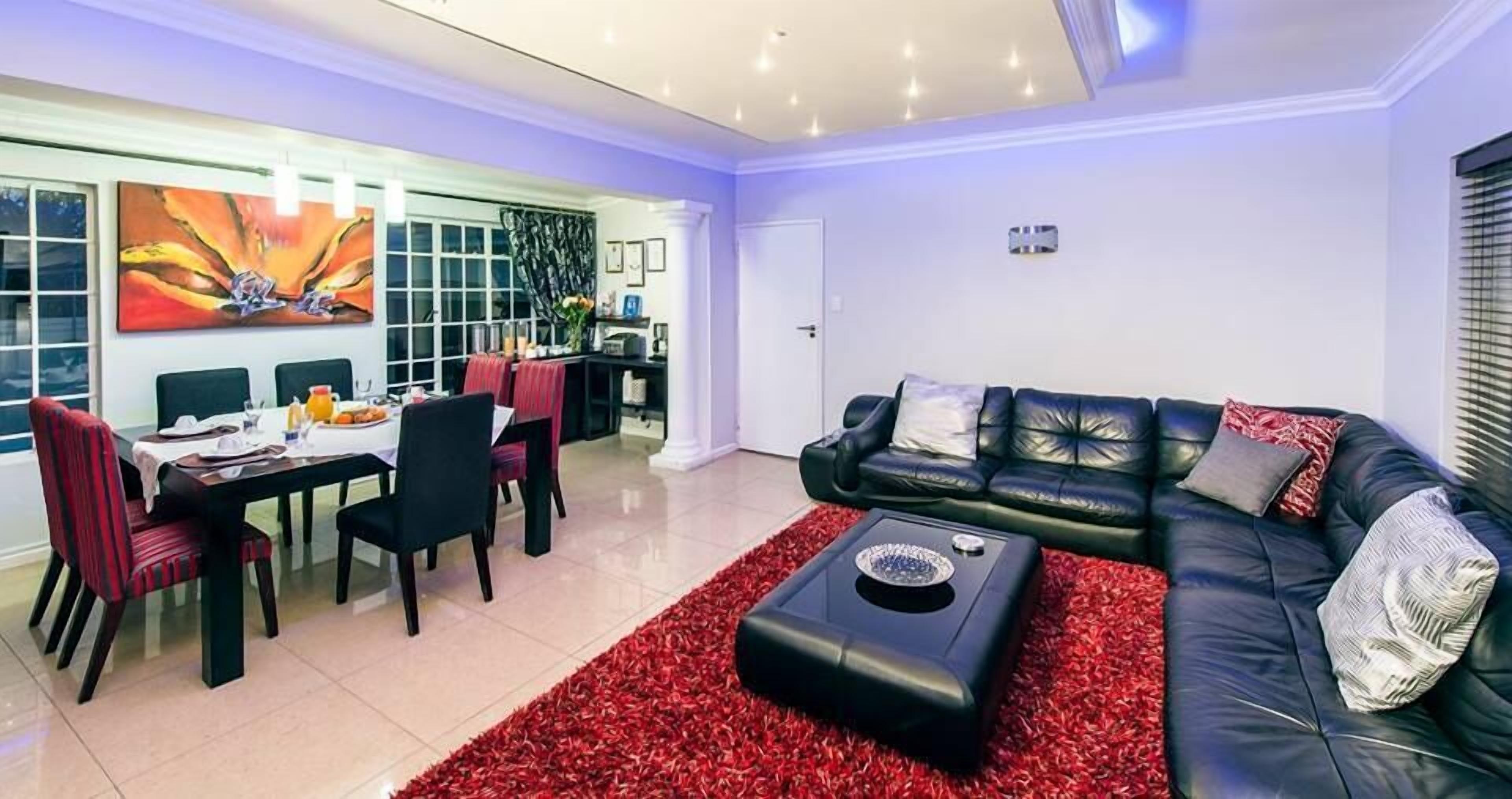 Uma sala de estar moderna com um sofá de couro preto, um tapete felpudo vermelho e uma área de jantar com mesa posta para dois.