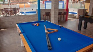 Uma mesa de bilhar com bolas e tacos, uma mesa de futebol de mesa, e área de estar externa com cadeiras e mesa.