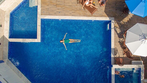 Vista aérea de uma piscina com uma pessoa boiando, cercada por espreguiçadeiras e guarda-sóis.