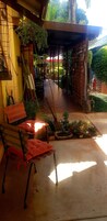 Uma área de estar coberta ao ar livre com cadeiras e mesas, cercada por plantas em vasos e um jardim.