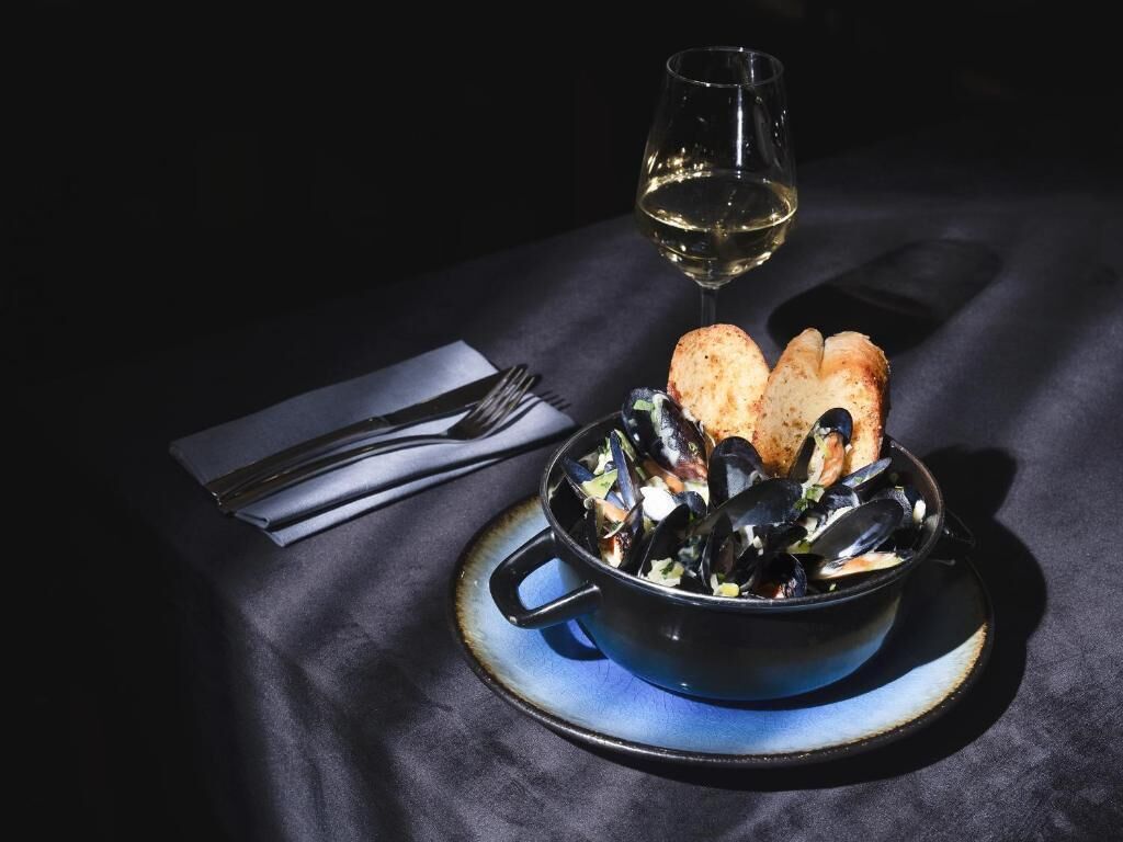 Uma tigela de mexilhões com pão numa travessa azul, um copo de vinho branco e um guardanapo dobrado sobre um tecido escuro.