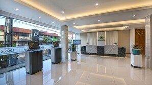 Uma área de recepção de hotel com design moderno, destacando um balcão de atendimento, uma tela digital e uma grande janela com vista para a rua.