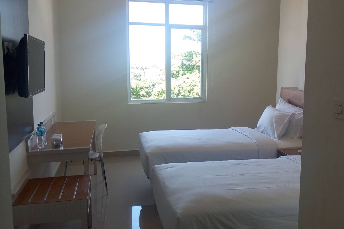 Quarto de hotel com duas camas, uma escrivaninha com cadeira, uma janela e uma televisão na parede.