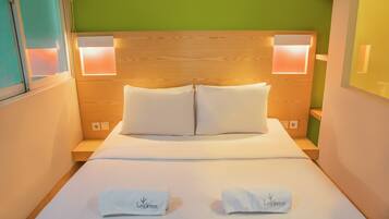 Quarto de hotel com cabeceira de madeira, duas luminárias de apoio, cama com roupa branca e duas toalhas com o nome do hotel.