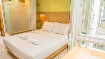 Quarto de hotel com cama, cabeceira de madeira, box de vidro para chuveiro, mesa de cabeceira e janela com vista para o verde.
