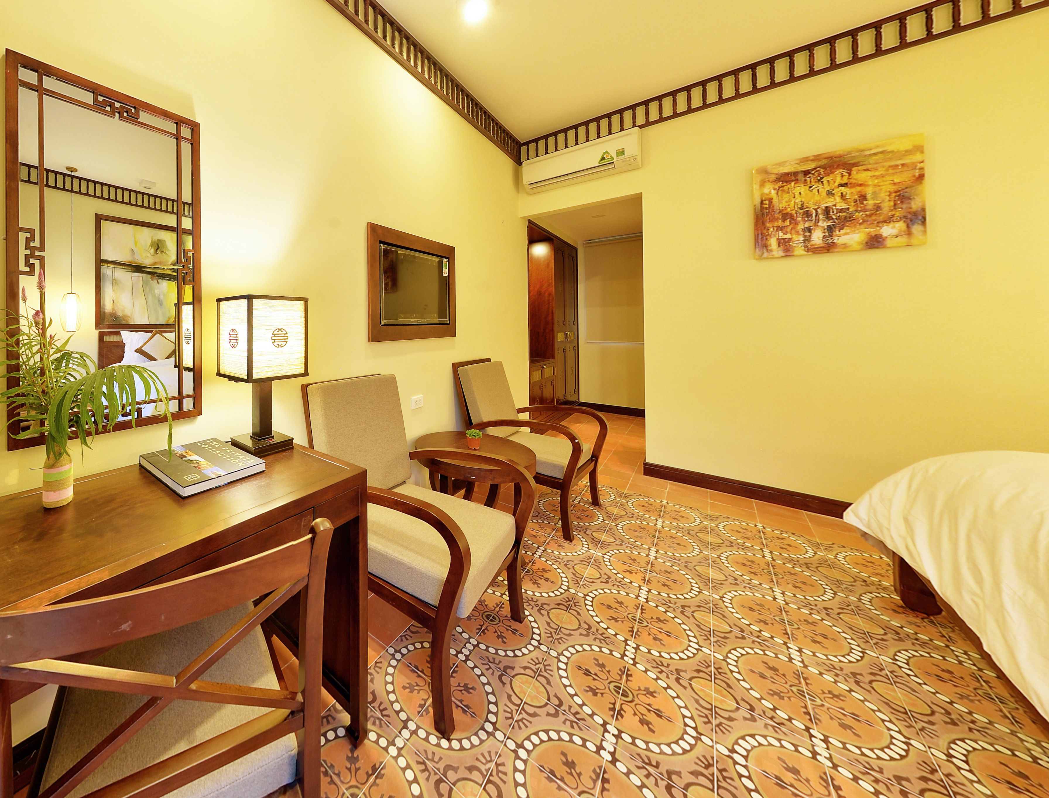 Quarto de hotel com escrivaninha de madeira, duas cadeiras, uma cama, uma mesinha de cabeceira com abajur, um vaso de planta e um quadro na parede.