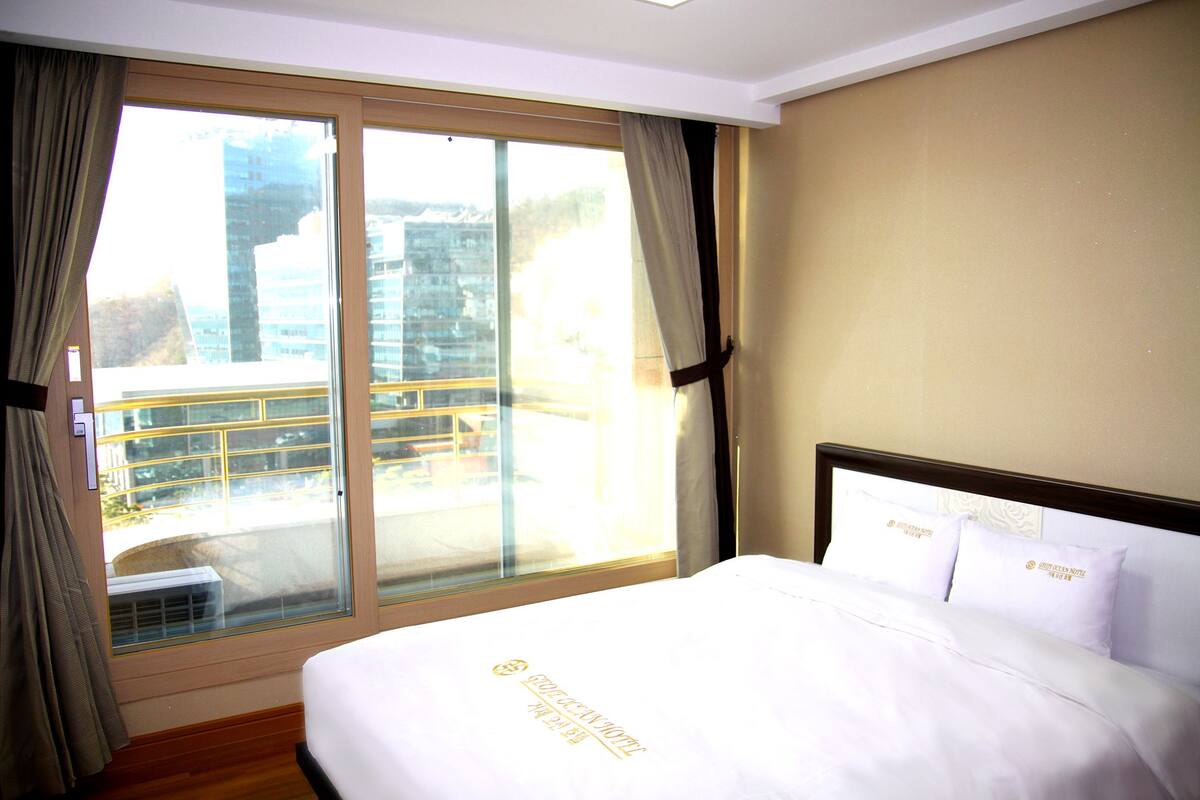Quarto de hotel com uma cama grande, janela com cortinas e vista para edifícios.