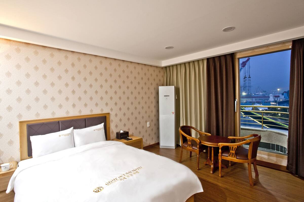 Quarto de hotel com cama, mesa de cabeceira, mesa com cadeiras, janela com vista para o horizonte urbano e parede com papel de parede estampado.