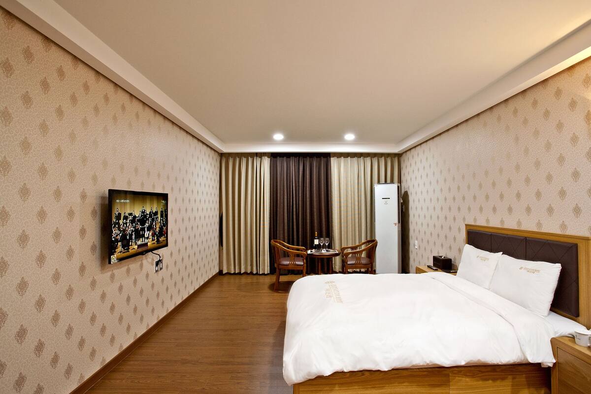 Quarto de hotel com uma cama grande, uma televisão na parede, uma mesa de jantar com cadeiras e um frigorífico.