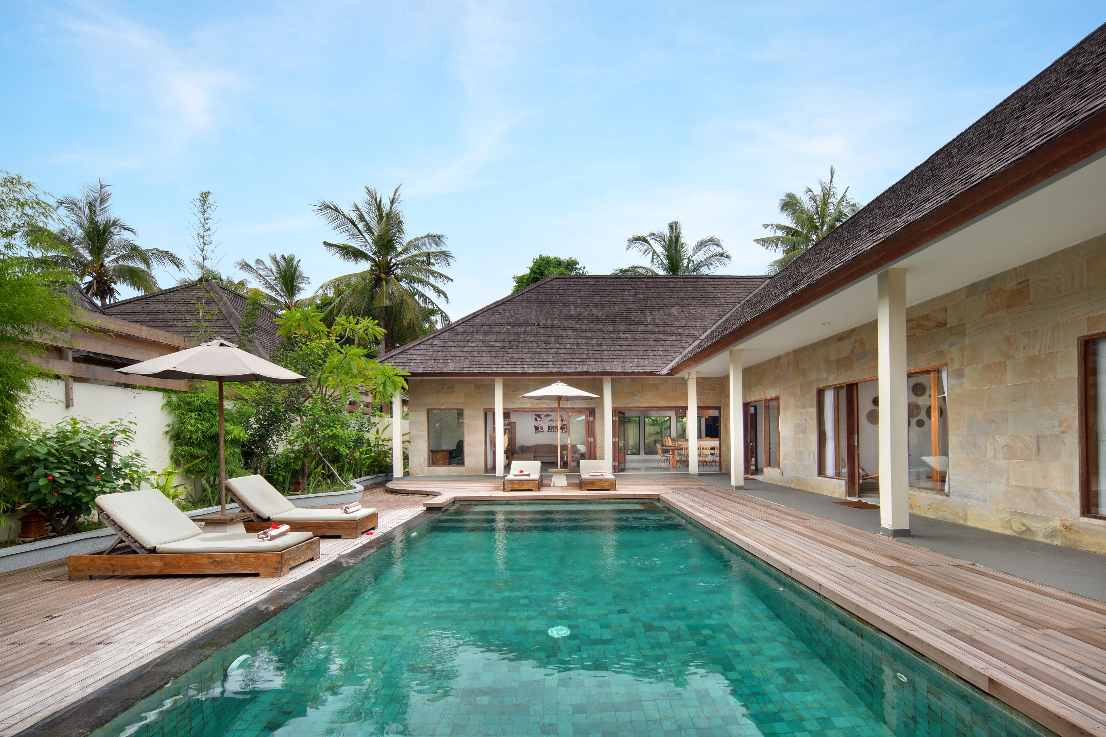 Piscina com espreguiçadeiras e guarda-sóis, cercada por um deck de madeira e palmeiras.