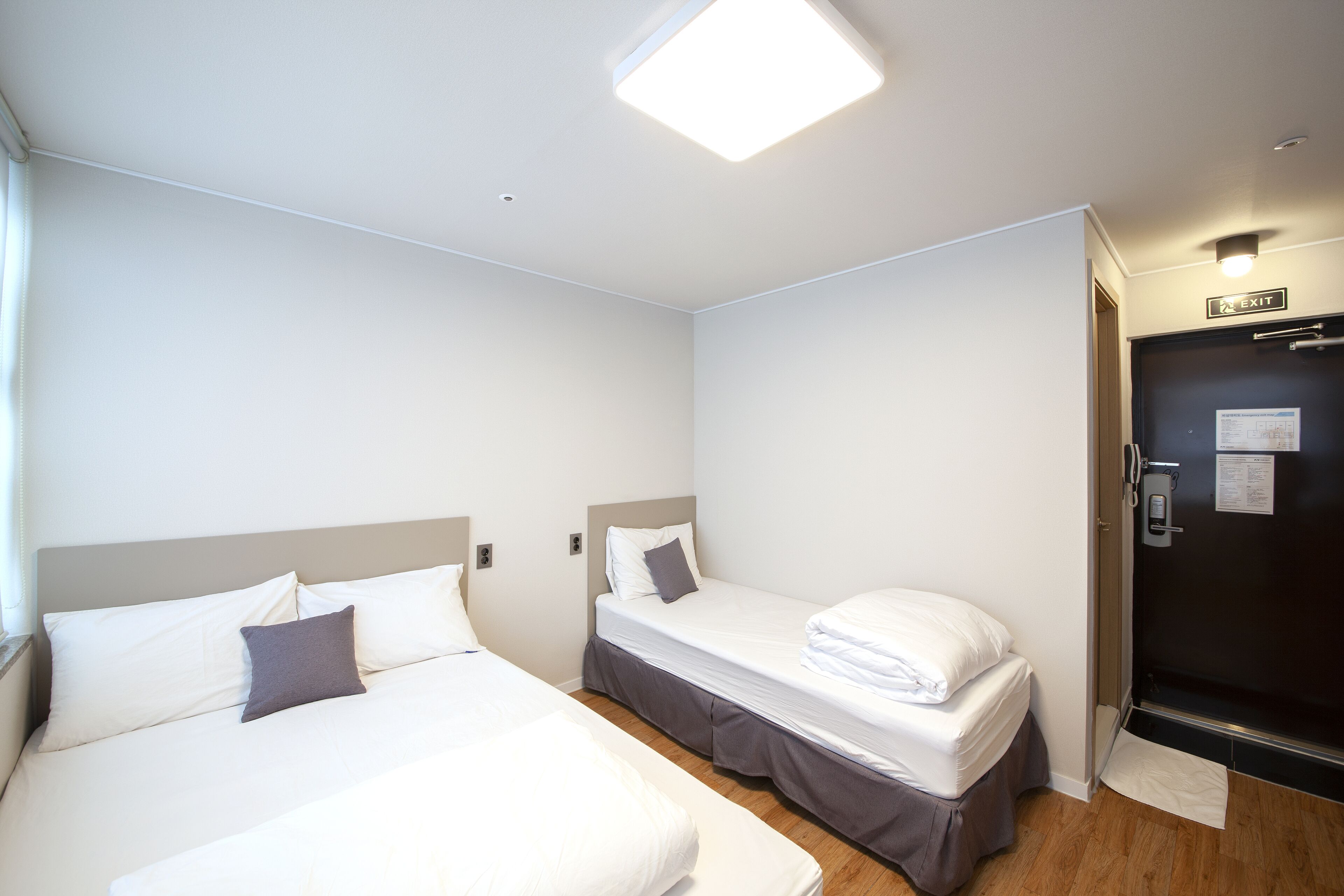 Quarto de hotel com duas camas, uma porta escura com sinal de saída e uma luz no teto.