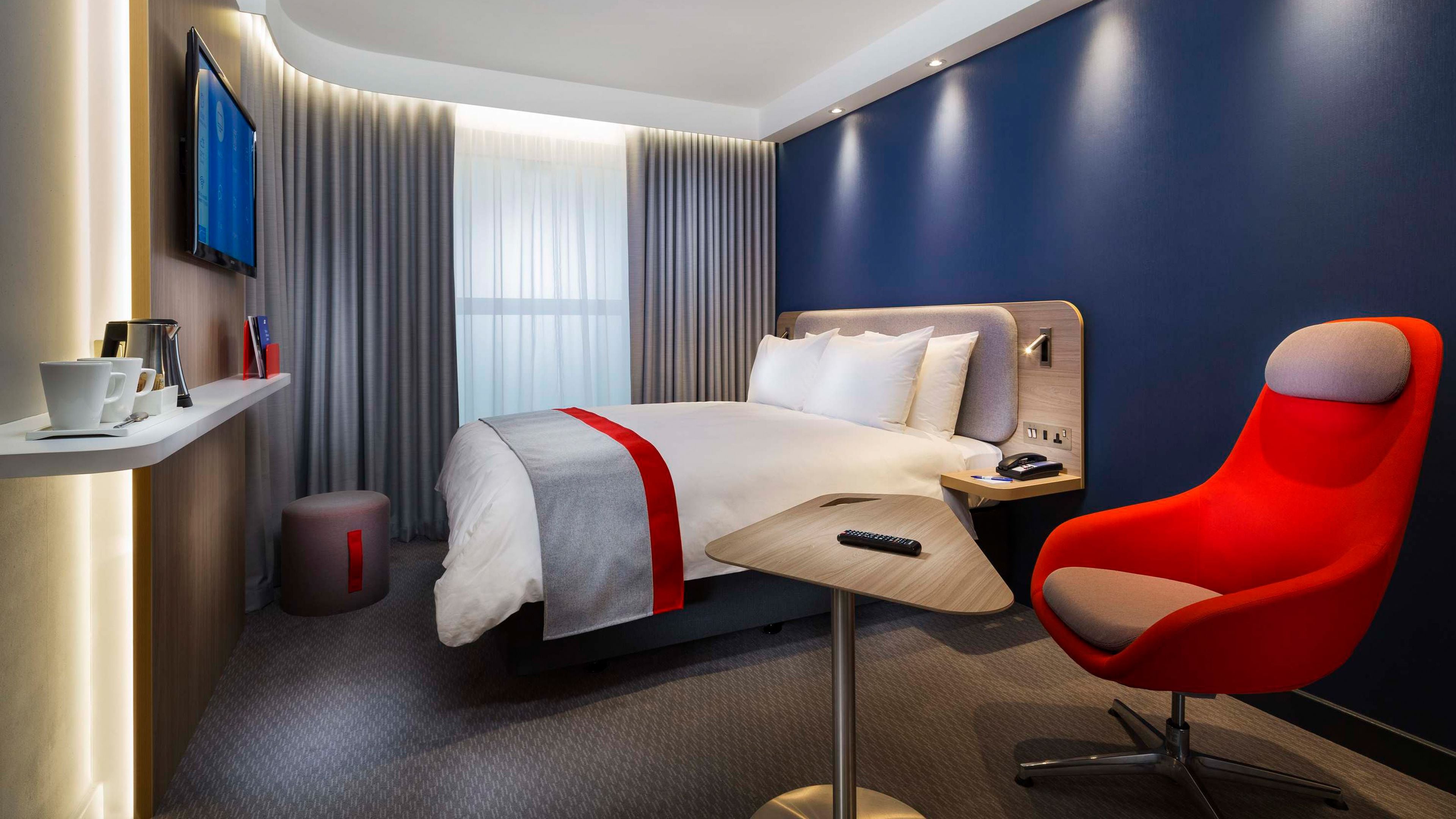 Quarto de hotel moderno com uma cama, uma cadeira vermelha, uma escrivaninha com um controle remoto, uma lixeira pequena e uma estante com xícaras e livros.