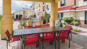 Um pátio externo com mesa de metal e cadeiras vermelhas, churrasqueira e vista para um edifício com paredes de tijolo e tons bege.