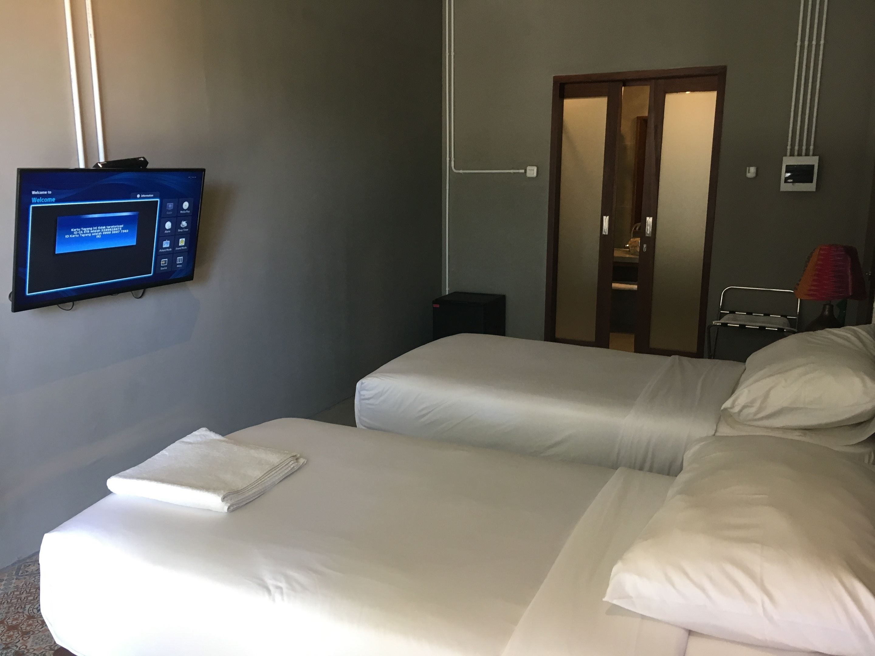 Quarto de hotel com duas camas, uma televisão fixada na parede, uma mesinha de cabeceira e uma porta que dá para outro quarto.