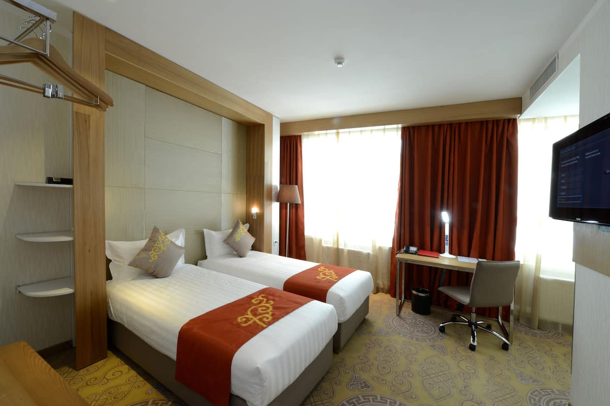 Quarto de hotel com duas camas, uma escrivaninha, uma cadeira, uma televisão e uma janela com cortinas.