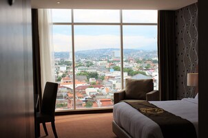 Quarto de hotel com uma grande janela, cama, poltrona e vista para a paisagem urbana.