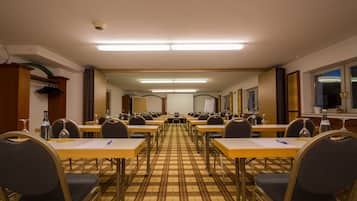 Sala de conferências com fileiras de mesas e cadeiras, equipada com garrafas de água e canetas.