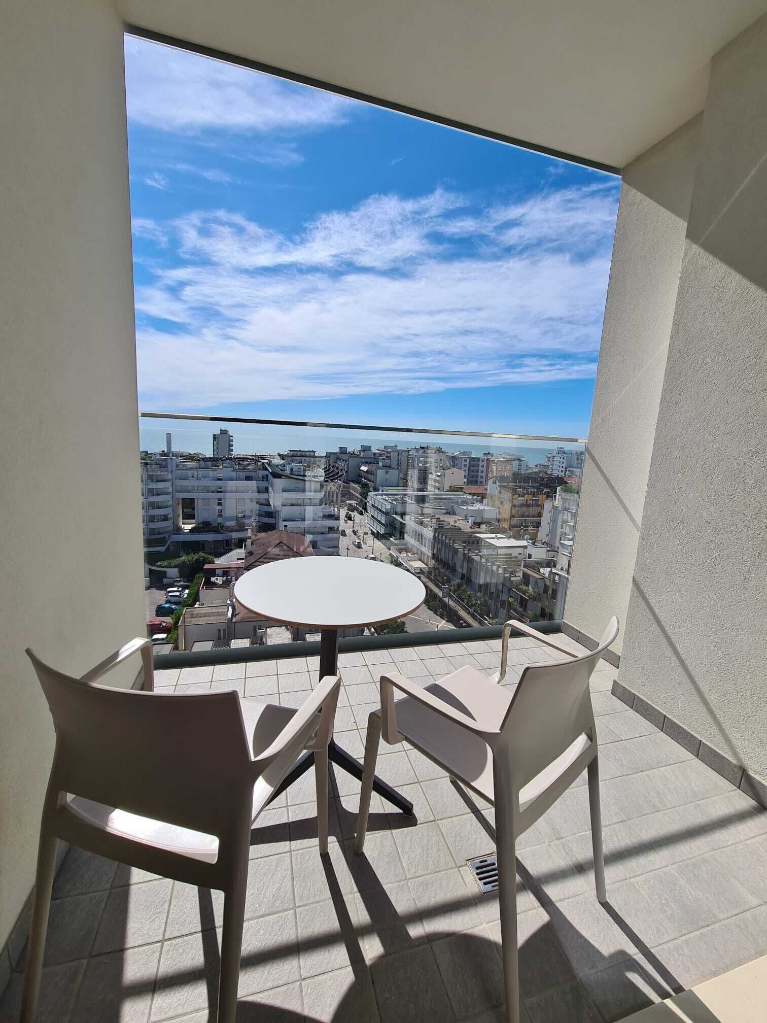 Uma varanda com uma mesa redonda e duas cadeiras, com vista para uma paisagem urbana com edifícios e um corpo d'água.