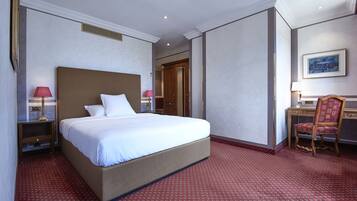 Quarto de hotel com uma cama grande, uma escrivaninha com cadeira, mesa de cabeceira e abajur.