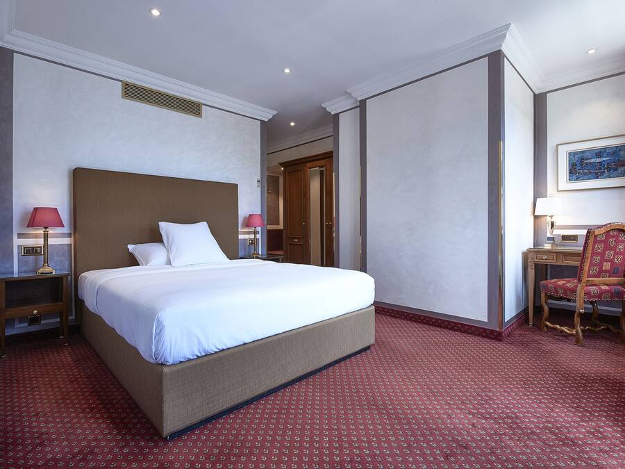 Quarto de hotel com uma cama grande, uma escrivaninha com cadeira, mesa de cabeceira e abajur.