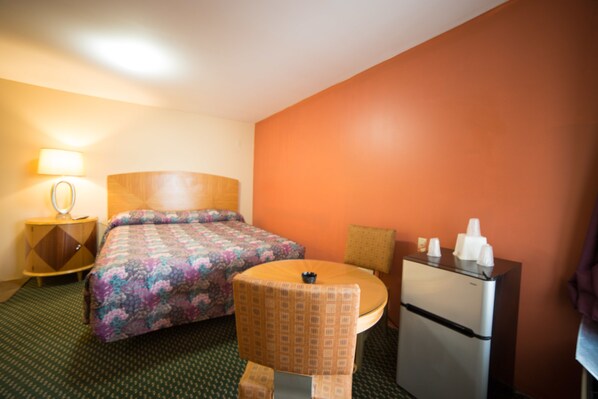 Quarto de hotel com cama, uma pequena mesa, cadeira, frigorífico e uma luminária de cabeceira.
