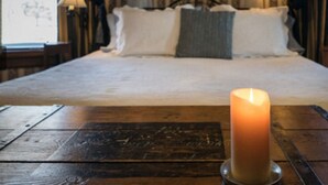 Uma cama com roupa de cama branca, uma vela acesa e uma mesa de madeira escura à frente.