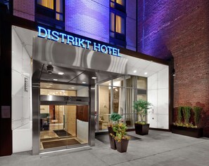 Exterior do District Hotel, com fachada moderna, entrada de vidro e plantas em vasos na área de acesso.