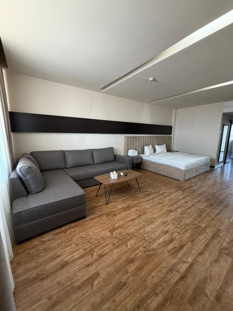 Quarto de hotel moderno com sofá modular, uma cama de solteiro, uma mesa pequena e piso de madeira.
