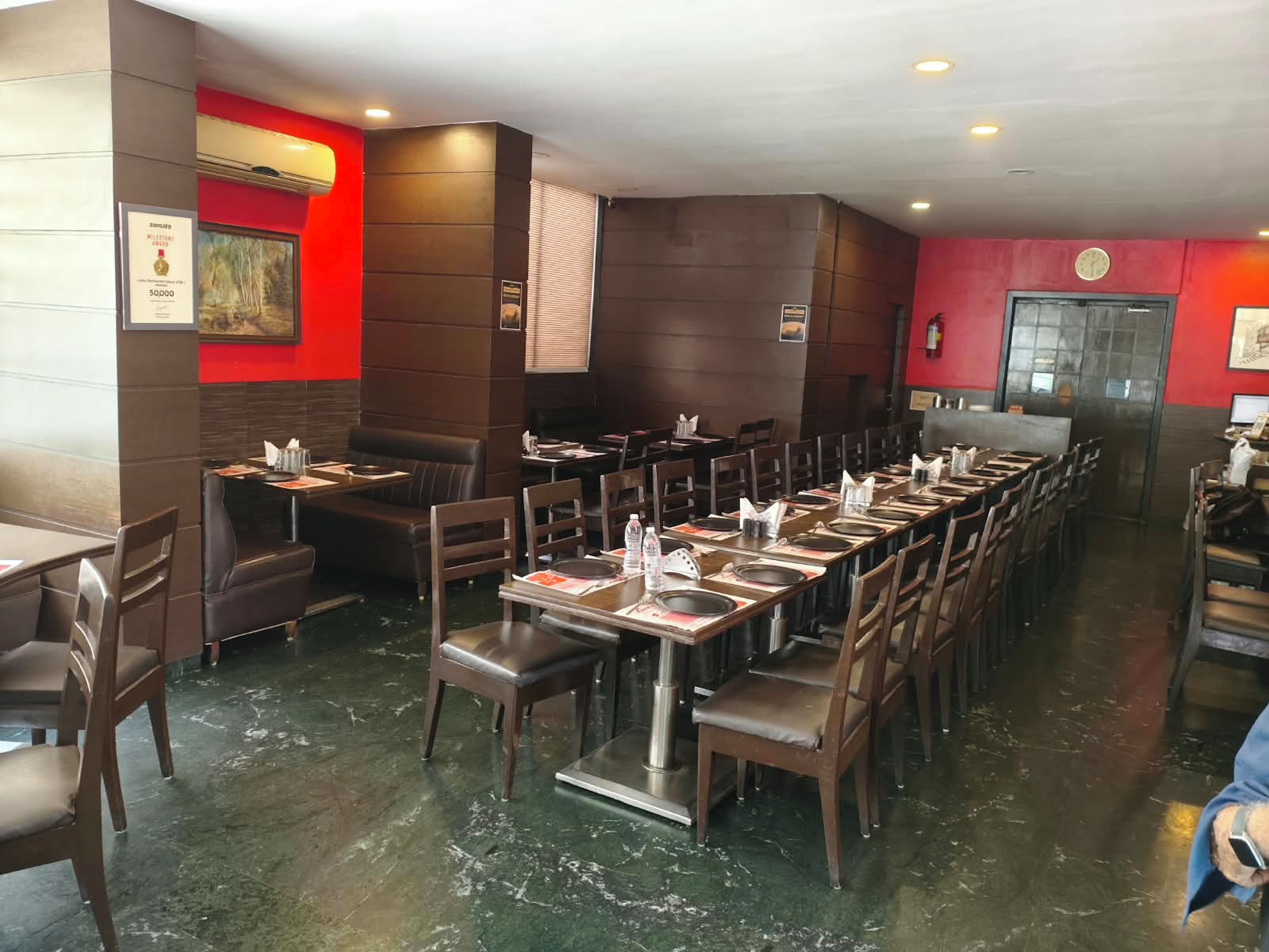 Um restaurante com mesas grandes e compartilhadas, cadeiras estofadas e uma parede vermelha decorativa, além de um relógio na parede.