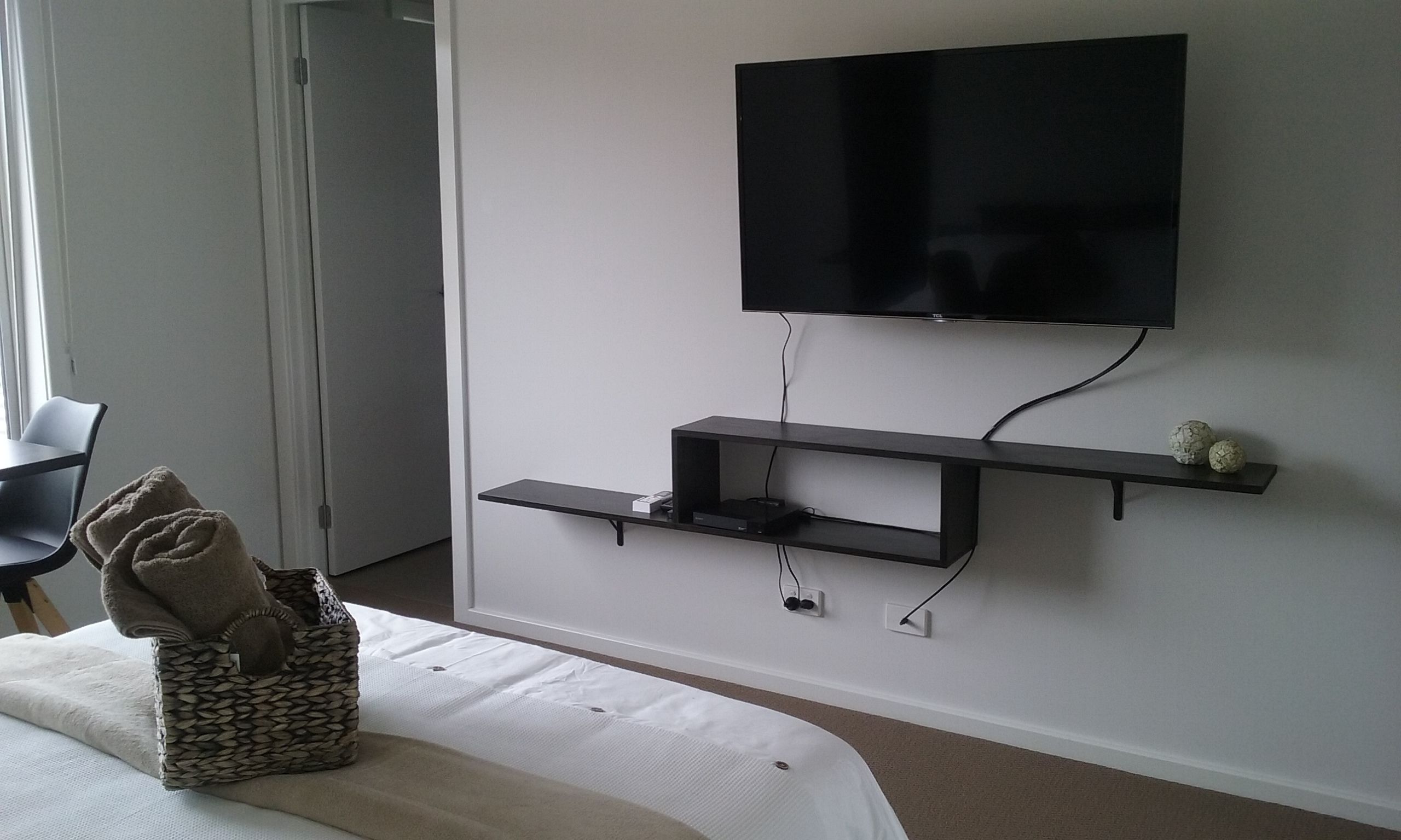 Um quarto moderno com uma cama, uma TV fixada na parede, um pequeno móvel de prateleiras abaixo da TV e um cesto de toalhas sobre a cama.
