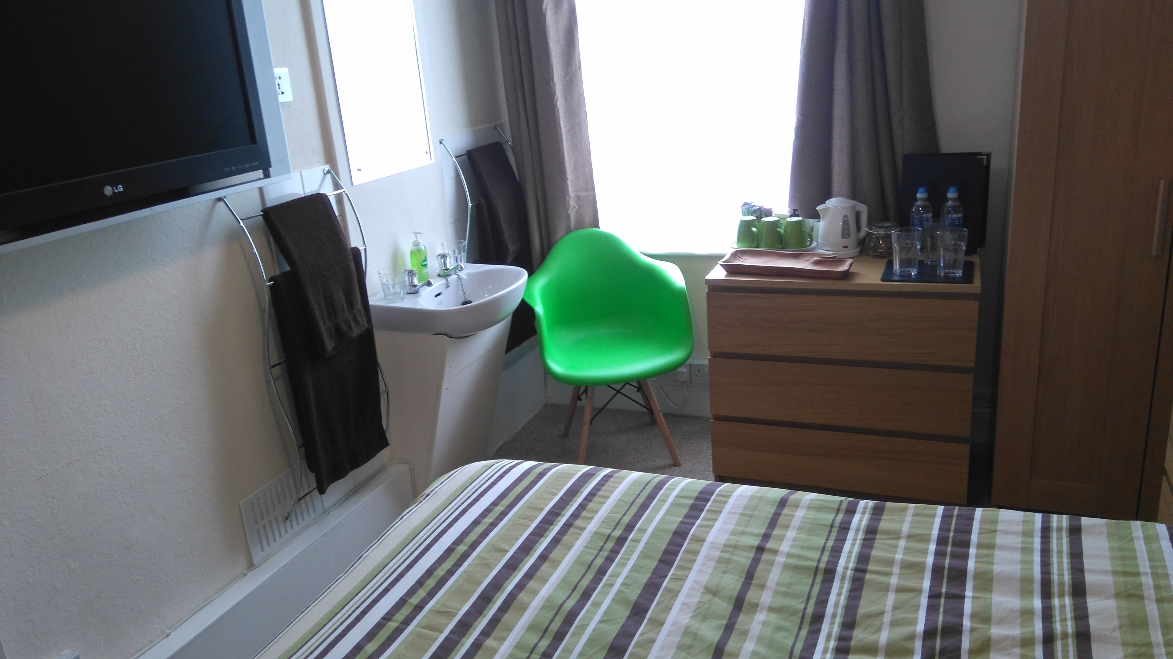 Quarto de hotel com uma cama, uma cadeira verde, cômoda de madeira, pia e uma TV de tela plana na parede.