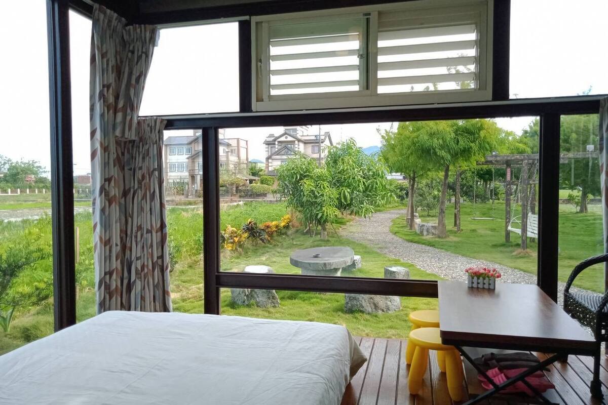Quarto com cama, uma mesinha e uma cadeira, com vista para um jardim com caminho e vegetação.