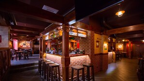 Uma área de bar com banquetas de madeira, parede de tijolos e balcão com diversos copos e garrafas.