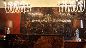 Balcão de recepção de hotel com bancada de mármore, duas luminárias de cristal e uma placa com a inscrição 'HOTEL EUROPA'.