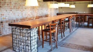 Um longo balcão de madeira com vários bancos e luminárias pendentes, em frente a uma parede de azulejos decorativos.