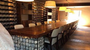 Uma longa mesa de madeira com cadeiras, cercada por prateleiras de vinho, em um ambiente com pouca luz.