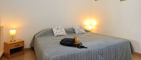 Cama bem arrumada com colcha cinza, dois travesseiros estampados, uma mesinha de cabeceira com abajur e uma bolsa de mão na cama.