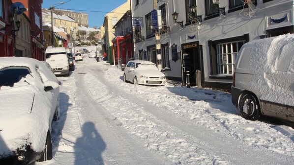 Rua coberta de neve com carros estacionados e edifícios, incluindo uma placa visível de um hotel.